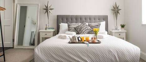 Cama bem arrumada com cabeceira cinzenta, roupa de cama branca e almofadas decorativas. Há uma bandeja com bule, xícaras e um vaso de flores sobre a cama.