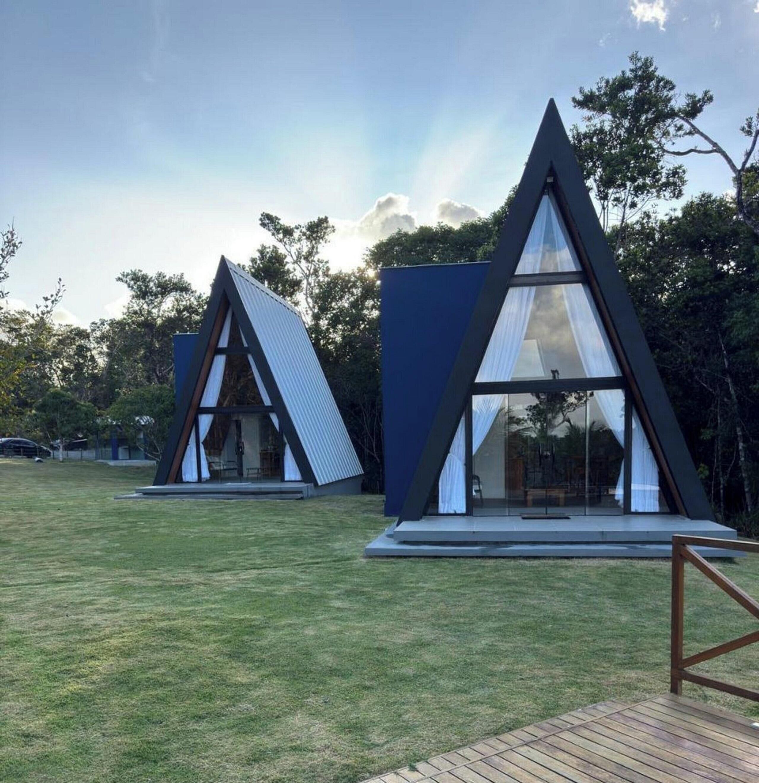 Duas cabanas modernas em formato de A, com grandes janelas de vidro, localizadas numa área gramada com árvores ao fundo.