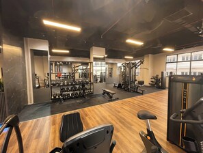 Ginásio com diversos equipamentos de exercício, piso de madeira e grandes janelas.