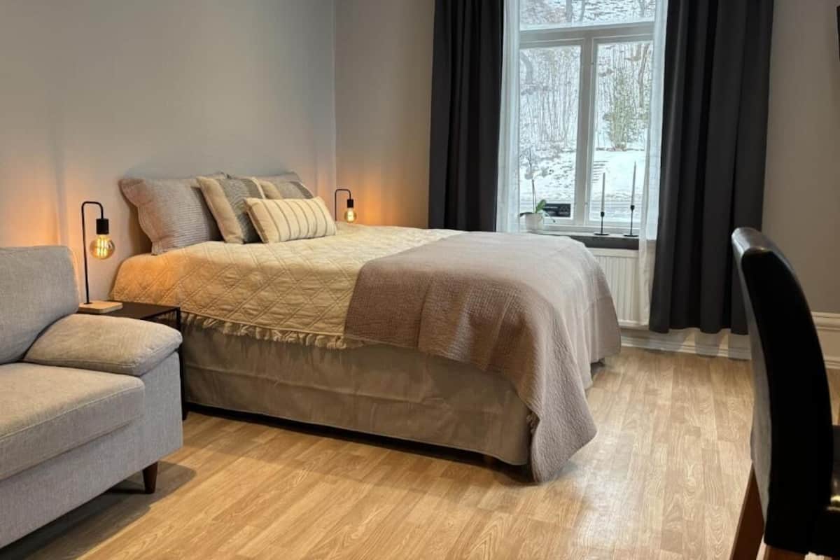 Um quarto com cama, uma cadeira, mesa de cabeceira e janela com cortinas.