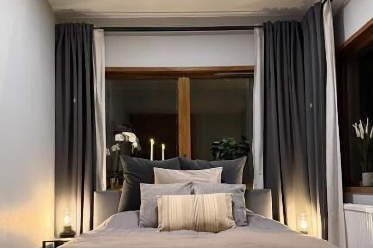 Quarto com cama, um banco, janela com cortinas e luminária no teto.