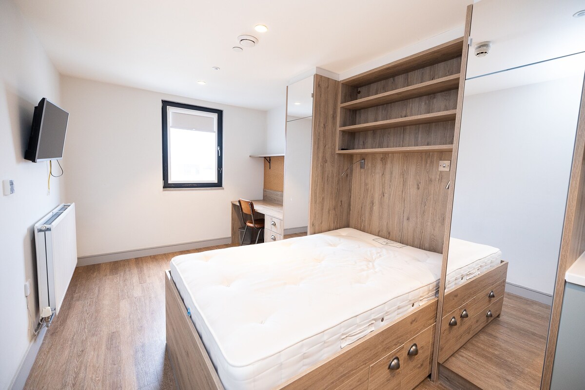 Quarto com cama, guarda-roupa de madeira, escrivaninha e televisão.