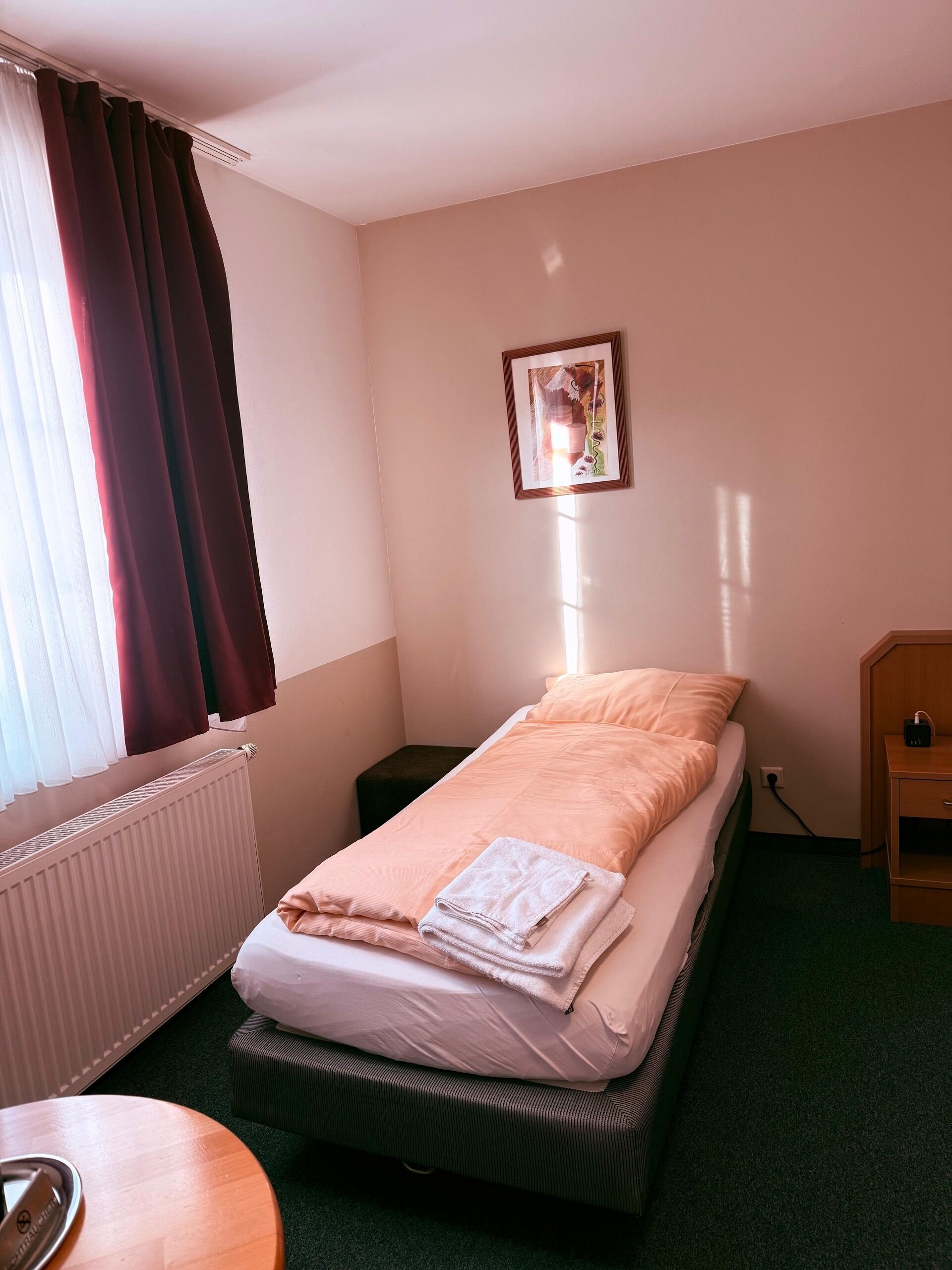Uma cama pequena e bem arrumada com um cobertor rosa e uma toalha branca dobrada, uma mesa de cabeceira de madeira com um abajur, um radiador e um quadro na parede.