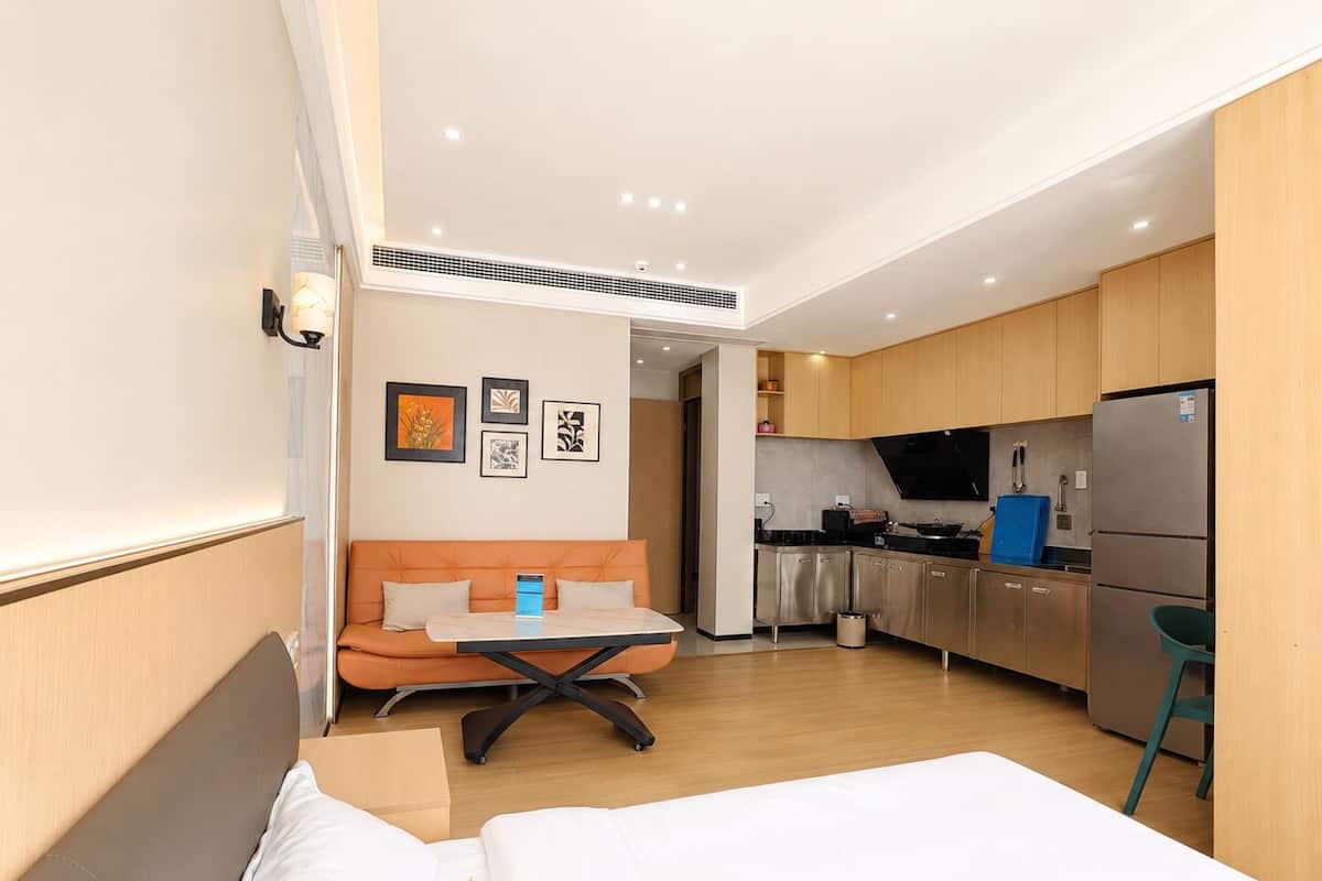 Quarto de hotel compacto com cama, uma mesinha, sofá, cozinha americana e banheiro visível através de uma porta aberta.