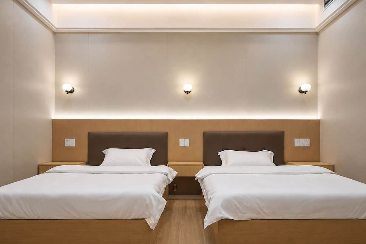 Quarto de hotel com duas camas, cabeceira de madeira, claraboia e iluminação embutida na parede.
