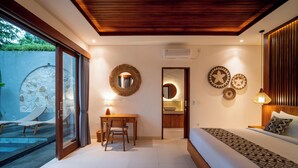 Quarto com cama, escrivaninha e banheiro pequeno. Apresenta uma parede de pedra e uma porta de vidro que dá para uma área de piscina.