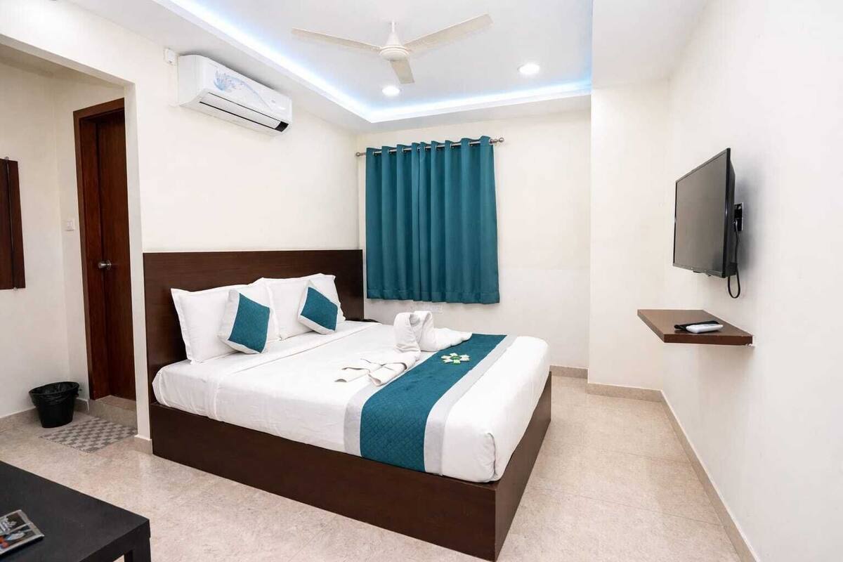 Quarto de hotel com cama, televisão, ventoinha no teto, ar condicionado e uma pequena mesa com controle remoto.