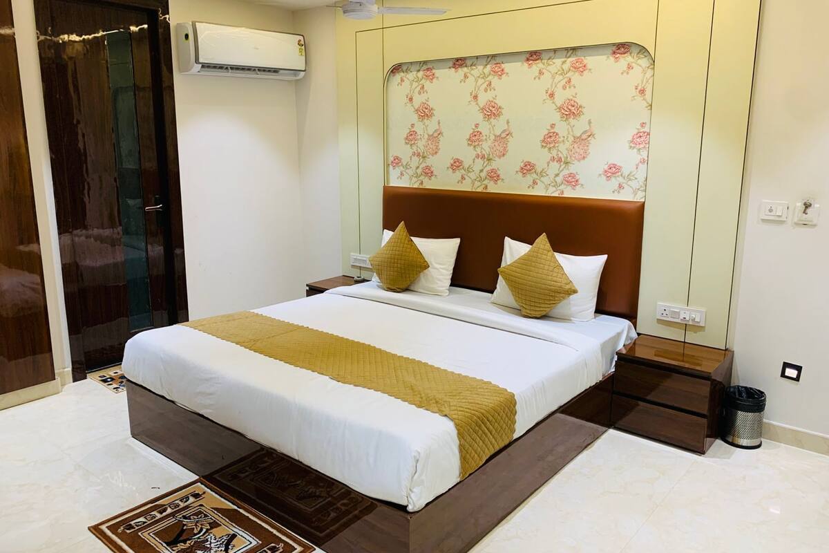 Quarto de hotel com cama, mesa de cabeceira, ar condicionado, ventilador de teto, porta de madeira e lixeira.