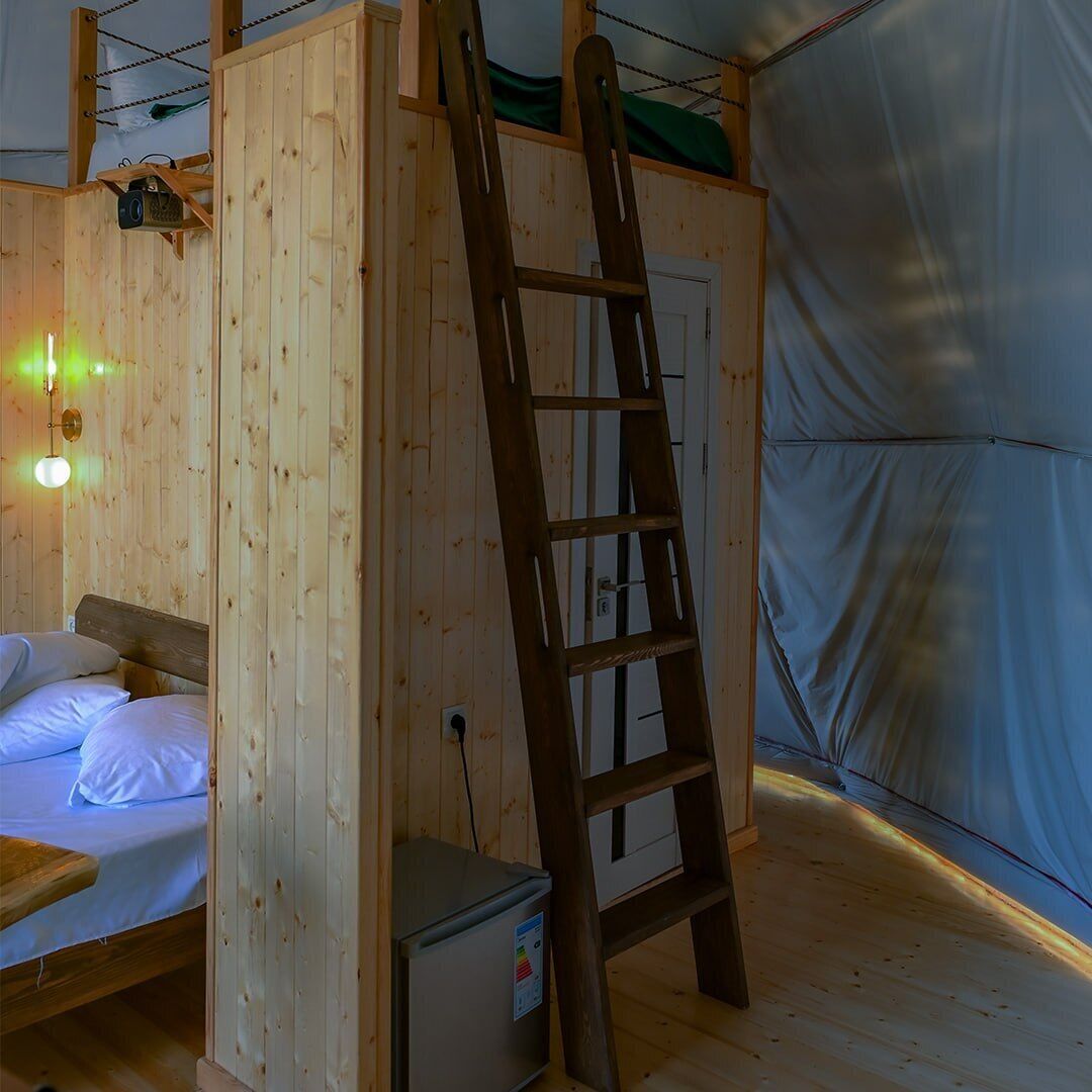 Uma cama beliche de madeira com escada, uma pequena escrivaninha e um alto-falante fixado na parede em um quarto tipo tenda.