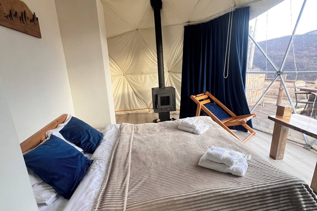 Quarto com tenda, uma cama, uma cadeira, uma mesa e um fogão a lenha.