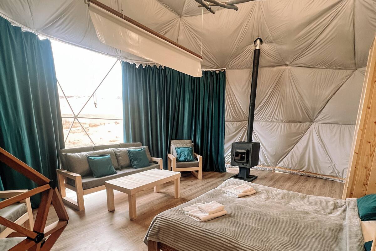 Acomodação em tenda com cama, duas cadeiras, uma mesa e um fogão a lenha.