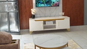 Uma sala de estar moderna com uma TV fixada numa parede de madeira, uma estante branca, uma mesa de centro redonda e um sofá castanho.