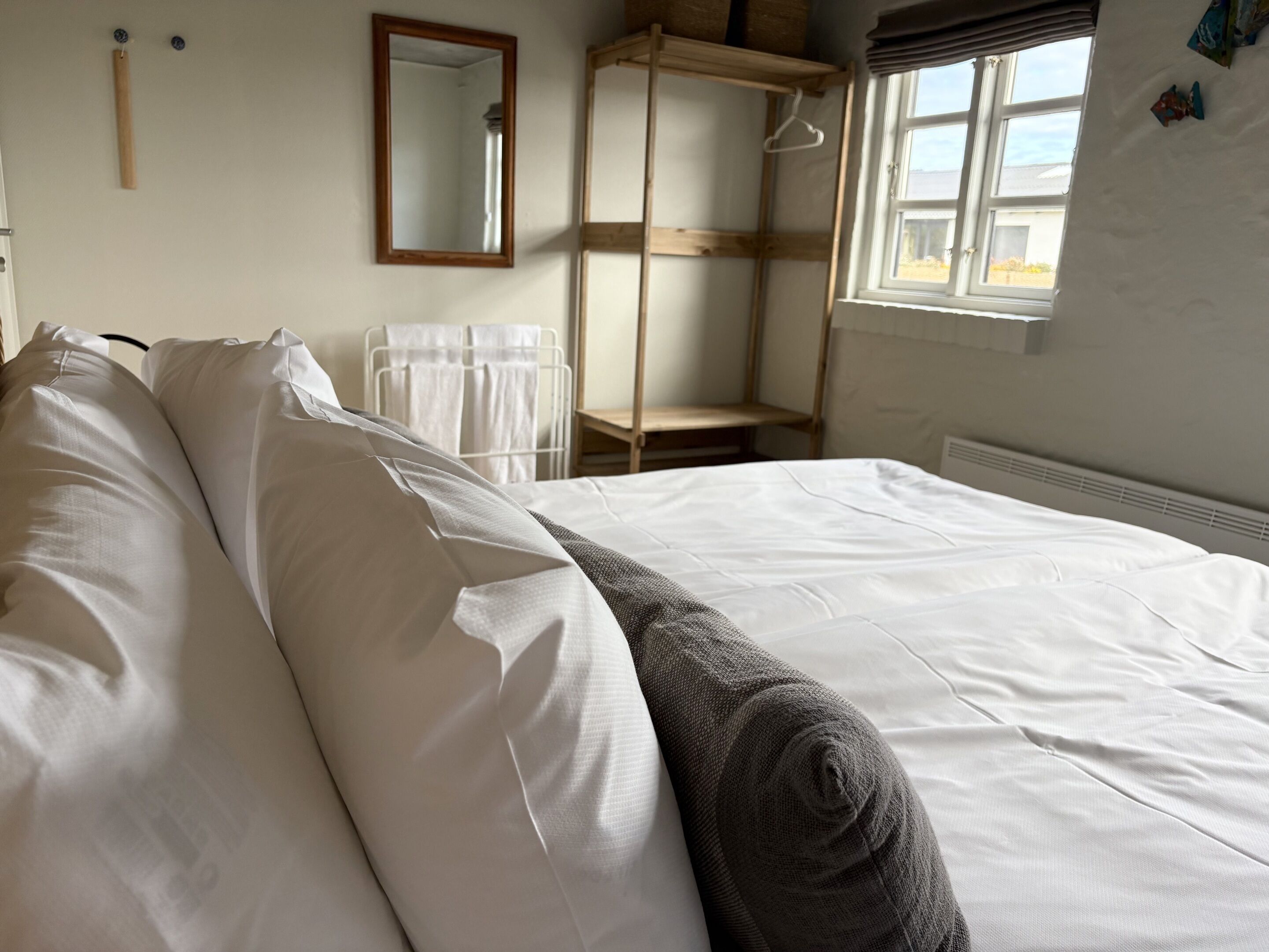 Uma cama com lençóis brancos, uma estante de madeira, um espelho e uma janela com luz do dia.