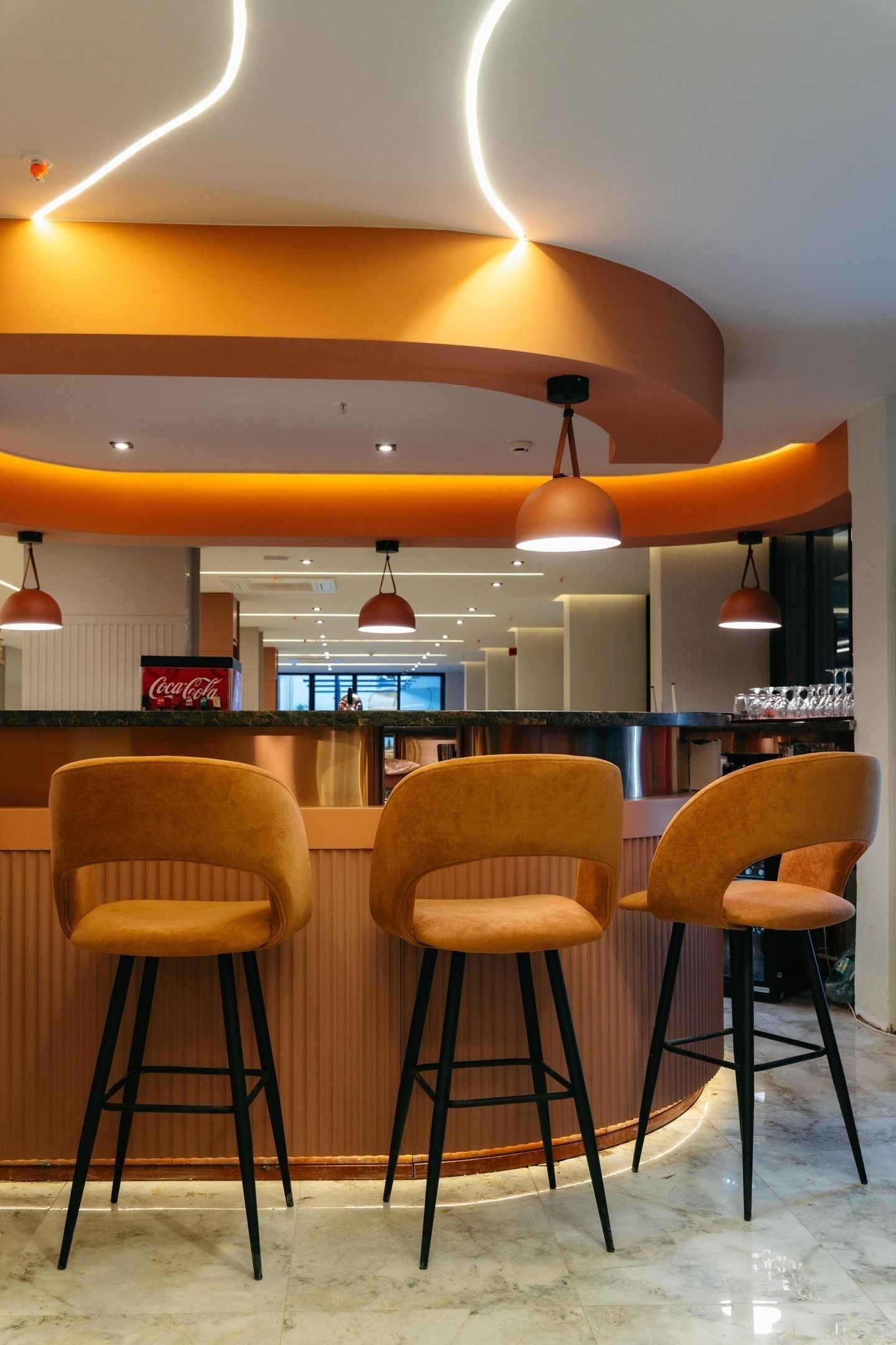 Uma área de bar com um letreiro da Coca-Cola, três banquetas e iluminação pendente.