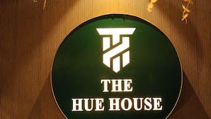 Um letreiro com o texto "THE HUE HOUSE" em um fundo verde, cercado por ripas de madeira verticais e alguma vegetação na parte inferior.