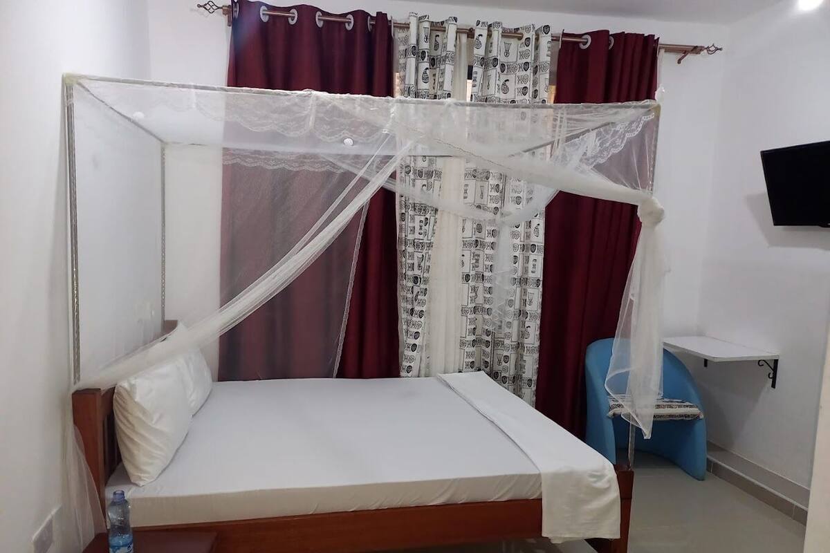 Uma cama de solteiro com dossel, uma mesa de cabeceira com um controle remoto e uma garrafa de água, uma cadeira azul pequena e uma TV fixada na parede.