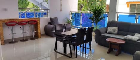 Uma sala de estar ampla com uma mesa de jantar, cadeiras, um sofá e uma pequena mesa de centro. Há três banquetas vermelhas e uma janela grande com vista para vegetação e uma estrutura azul lá fora.