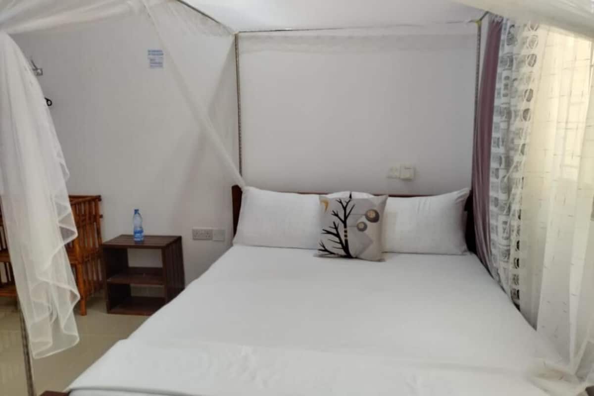 Uma cama de solteiro com dossel, uma mesinha de cabeceira com uma garrafa e um interruptor de luz fixado na parede.