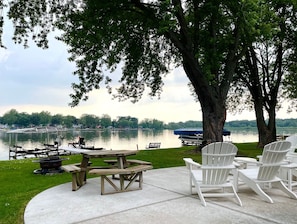 Um pátio à beira do lago com mesas de piquenique e cadeiras Adirondack, com vista para um lago calmo, com barcos e um píer ao fundo.