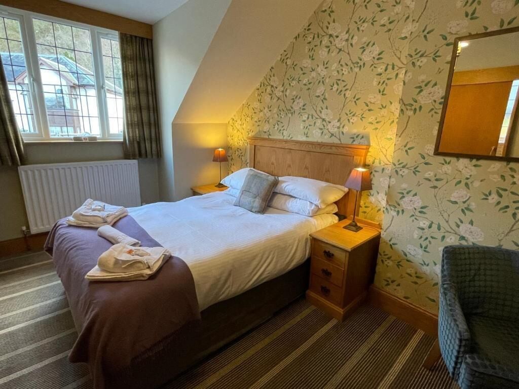 Quarto com cama, luminárias de cabeceira, mesa de cabeceira de madeira, uma cadeira e uma janela com cortinas.
