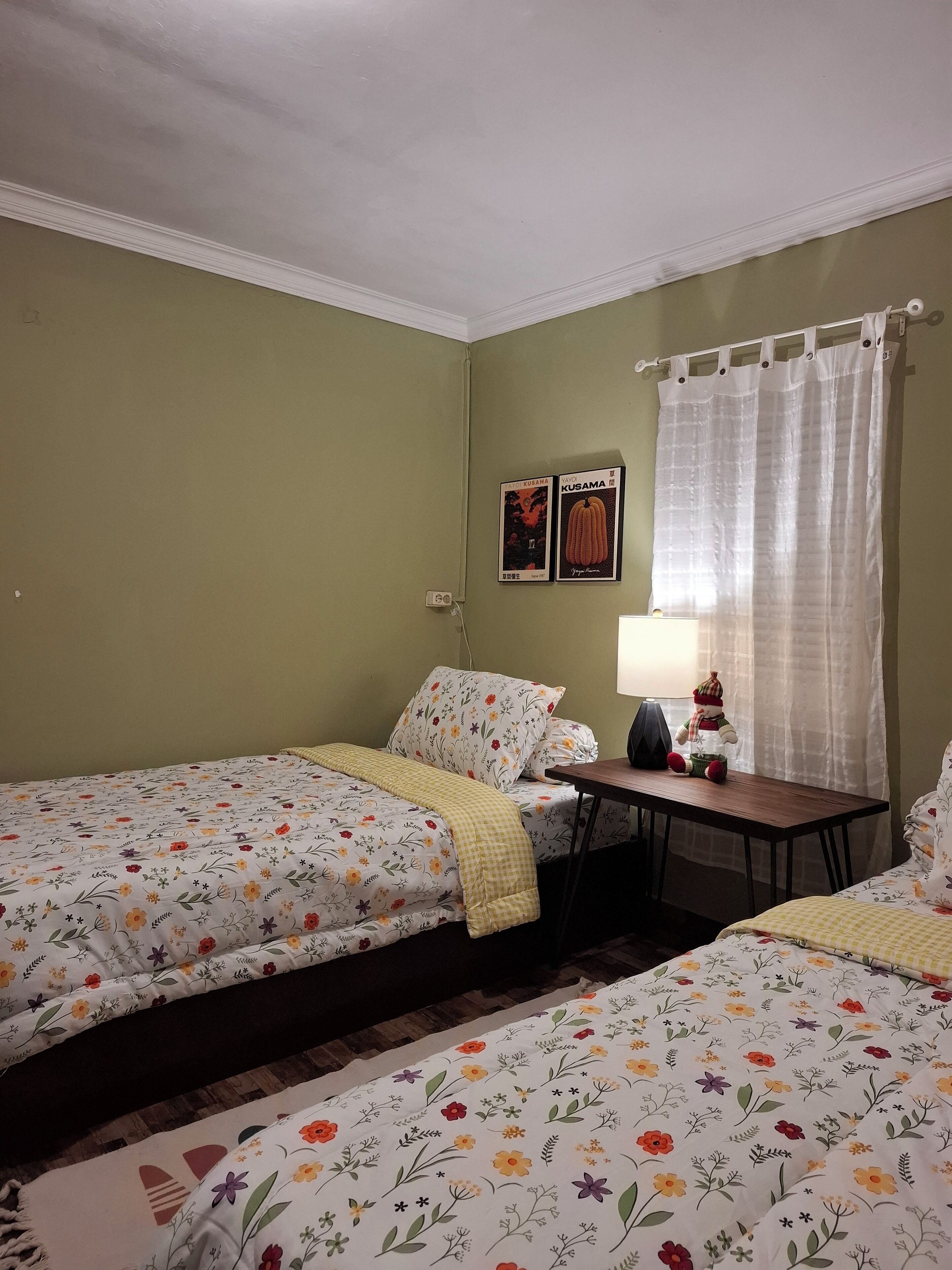 Quarto com duas camas, uma mesinha, uma luminária e dois cartazes na parede.