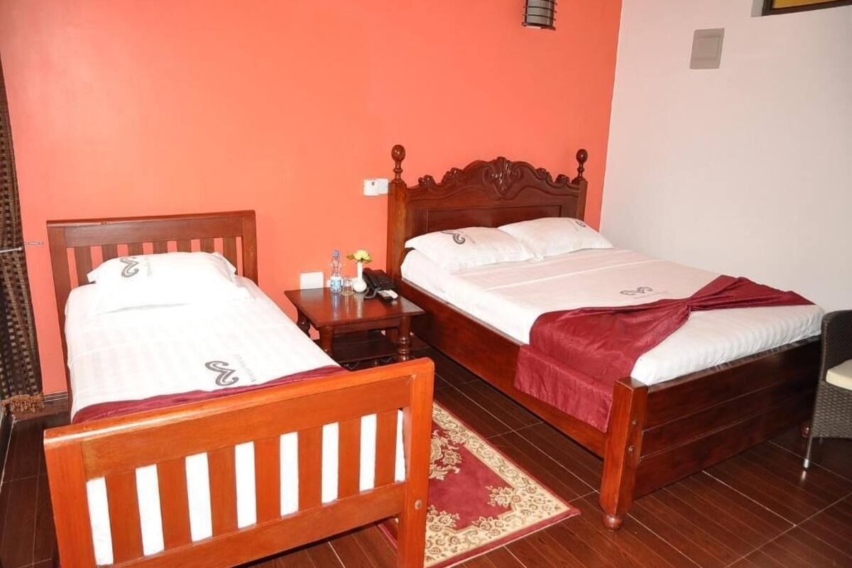 Quarto com duas camas de madeira, uma mesa pequena com um telefone e garrafas de água, e um tapete vermelho estampado.