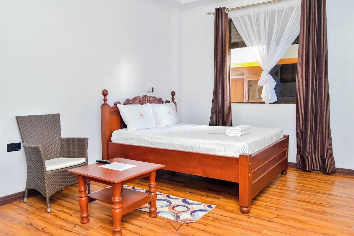 Quarto com cama de madeira, cadeira de vime, mesa pequena de madeira e janela com cortinas.