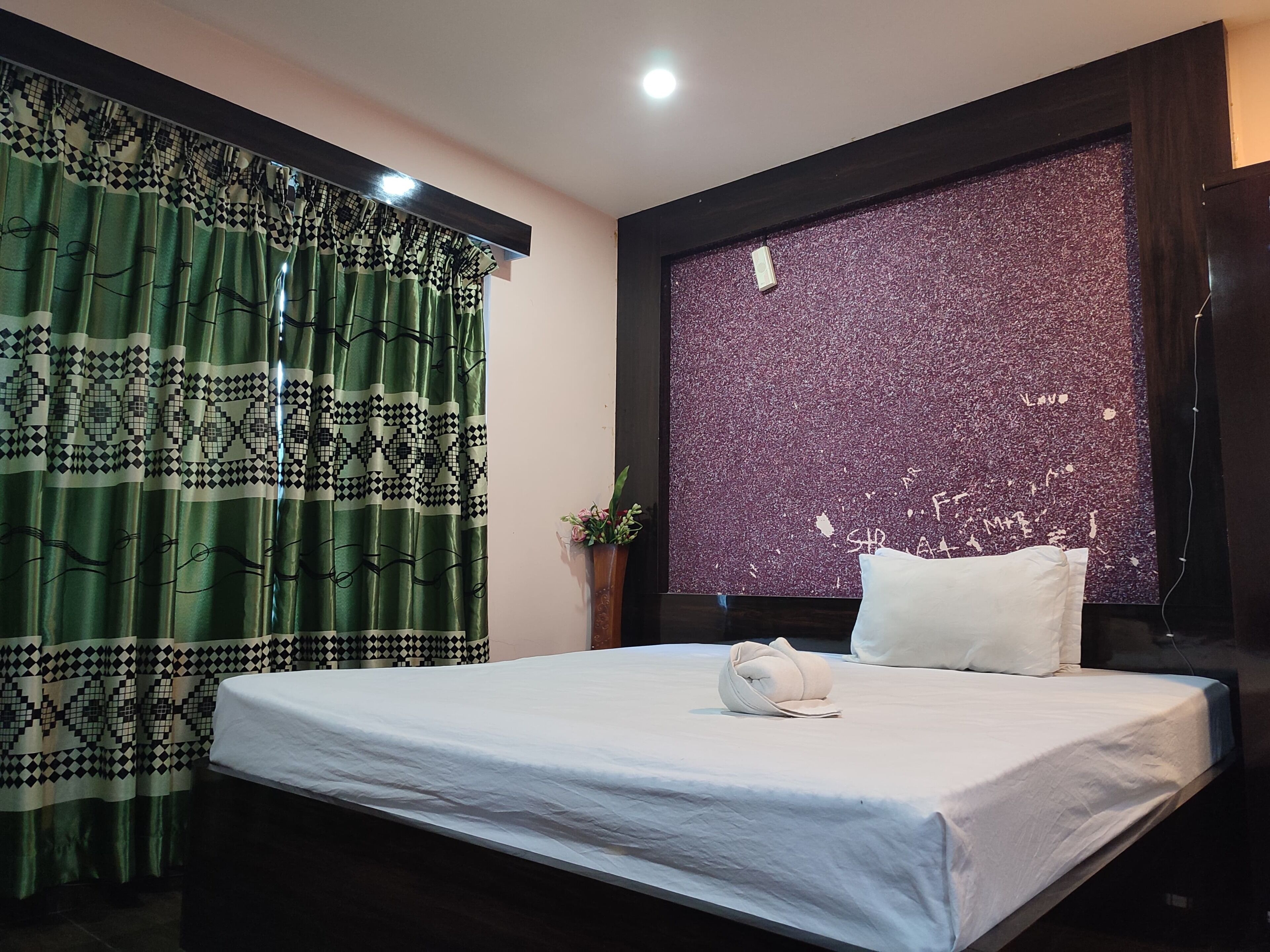 Uma cama bem arrumada com lençóis brancos e uma toalha dobrada, parede roxa com textura e cortina verde com padrão.