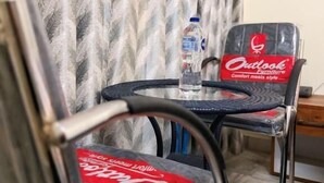 Uma cadeira com logótipo vermelho e branco, uma garrafa de água e uma cortina estampada num quarto.