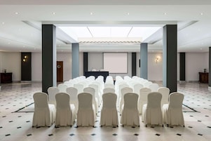 Uma sala de conferências com fileiras de cadeiras brancas, um púlpito e uma tela de projeção.