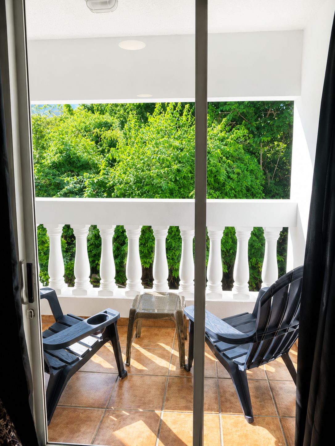 Uma varanda com grades brancas, duas cadeiras, uma mesa pequena e vista para a folhagem verde.