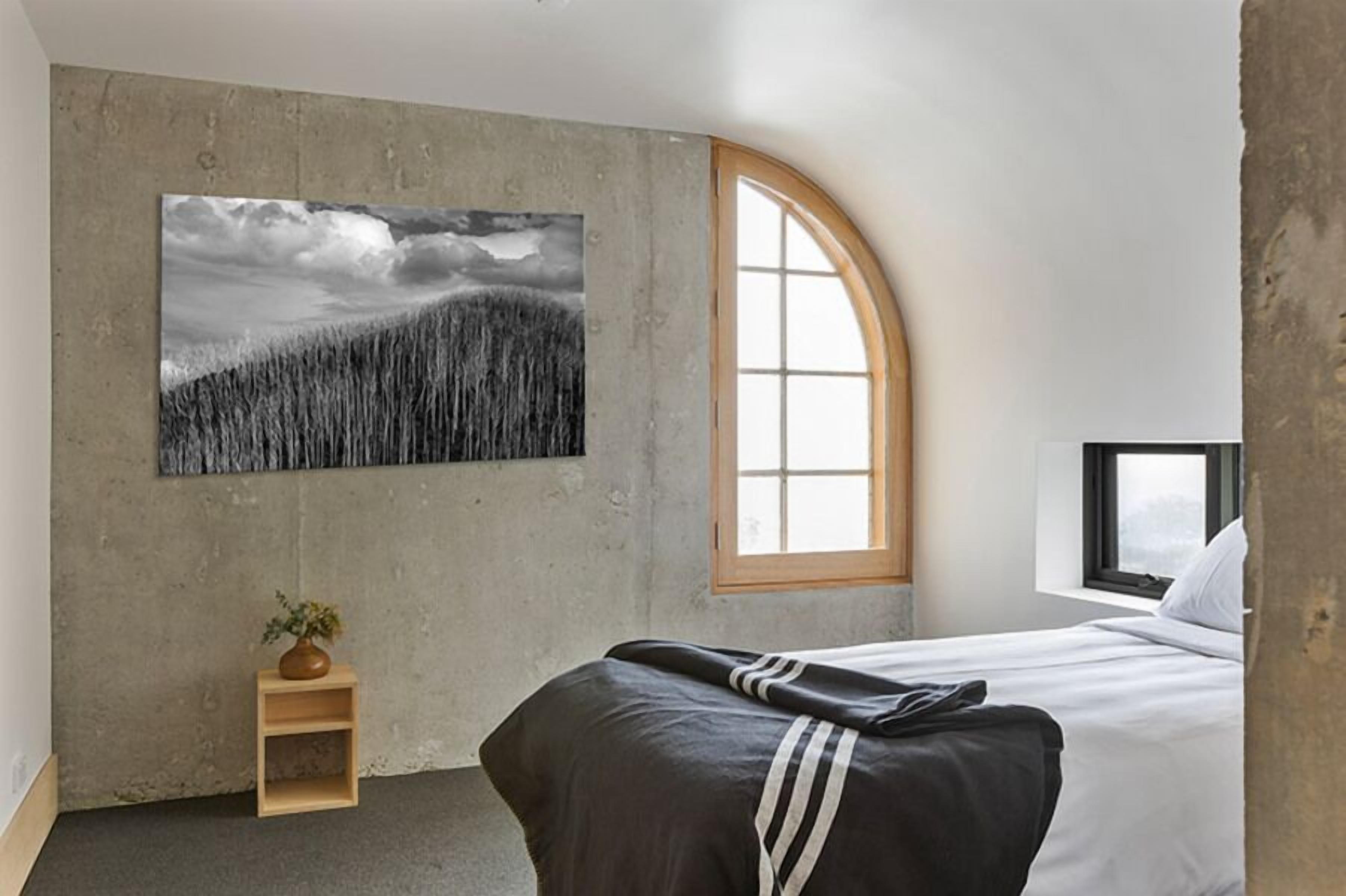 Quarto com uma grande pintura, um banquinho de madeira com uma planta, uma cama com manta listrada em preto e branco, e uma janela com vista para o céu.