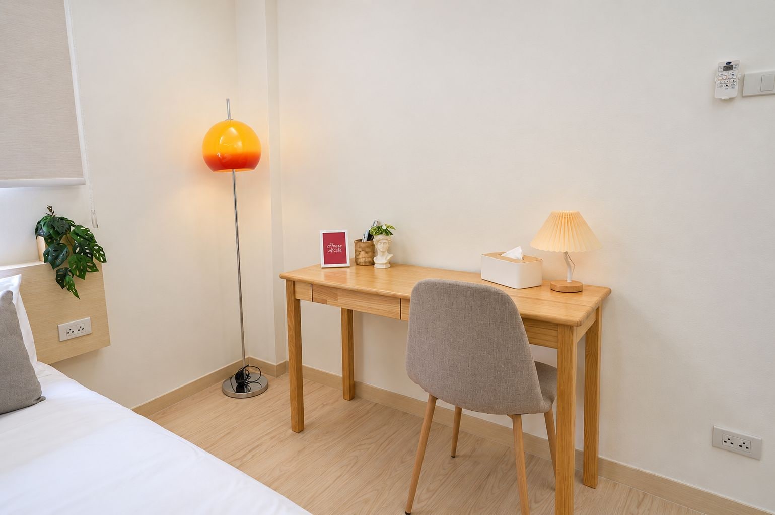 Quarto de hotel com escrivaninha de madeira, uma cadeira, mesa de cabeceira com abajur, planta na cabeceira e tomada na parede.