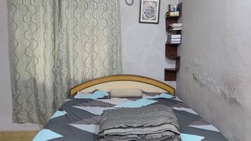 Quarto com cama, ar condicionado, cortina estampada, prateleira pequena com livros e um quadro na parede.