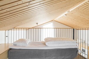 Quarto com teto inclinado, cama com roupa de cama branca e vista para o exterior através de uma janela.