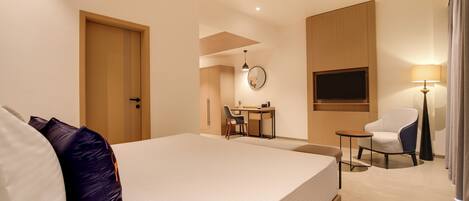 Quarto de hotel com uma cama grande, uma escrivaninha com cadeira, uma televisão e um abajur.
