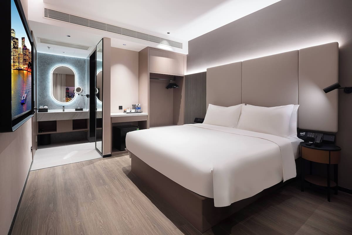 Quarto de hotel moderno com uma cama grande, uma mesinha de toilette com espelho, uma escrivaninha e uma televisão exibindo uma vista urbana.