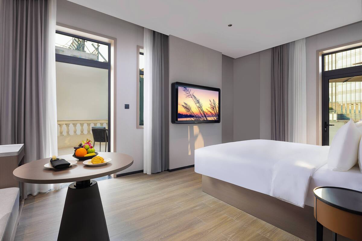 Quarto de hotel com cama, mesa de jantar com café da manhã, televisão na parede e janelas amplas.