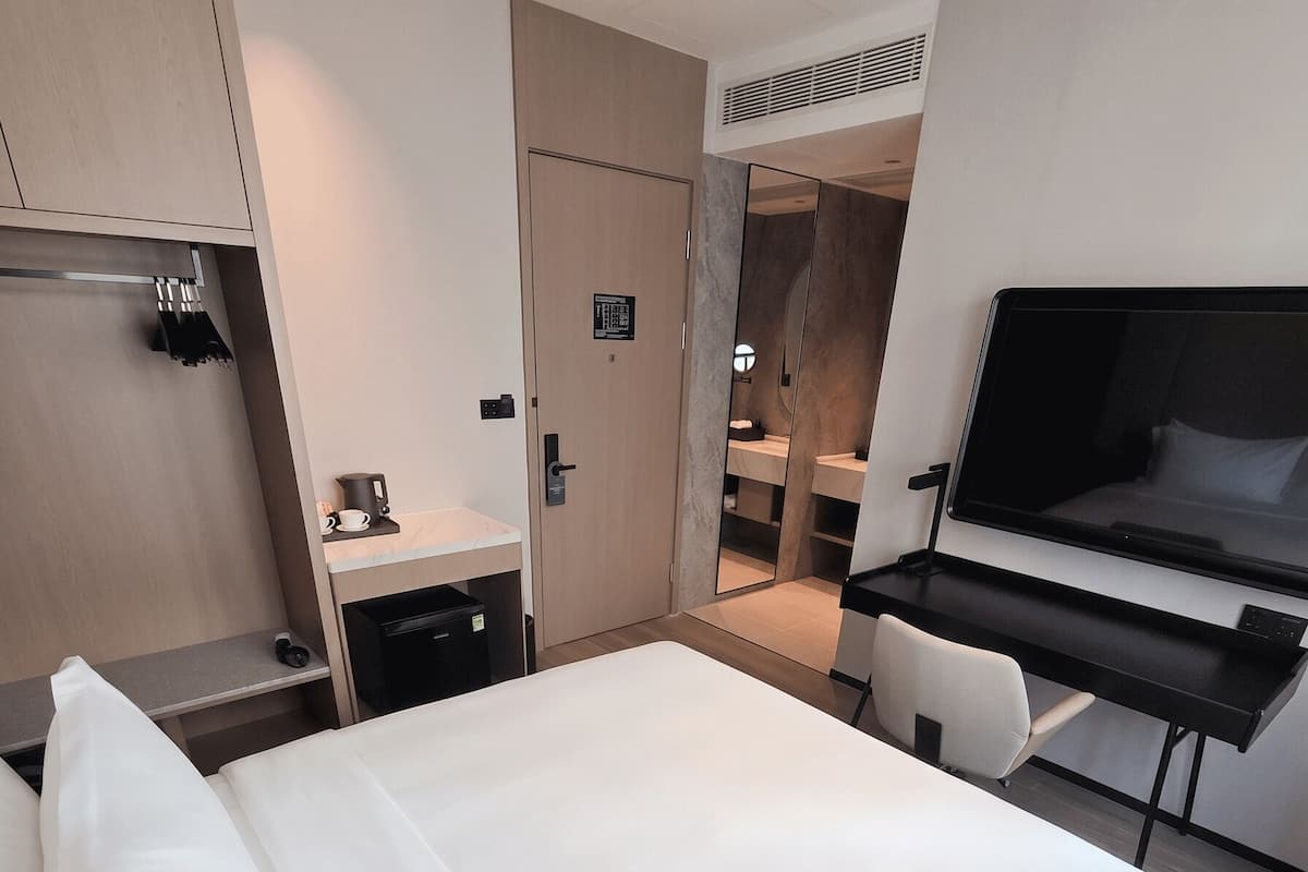 Quarto de hotel com uma cama, uma escrivaninha com televisão, um banheiro pequeno e uma porta com placa.