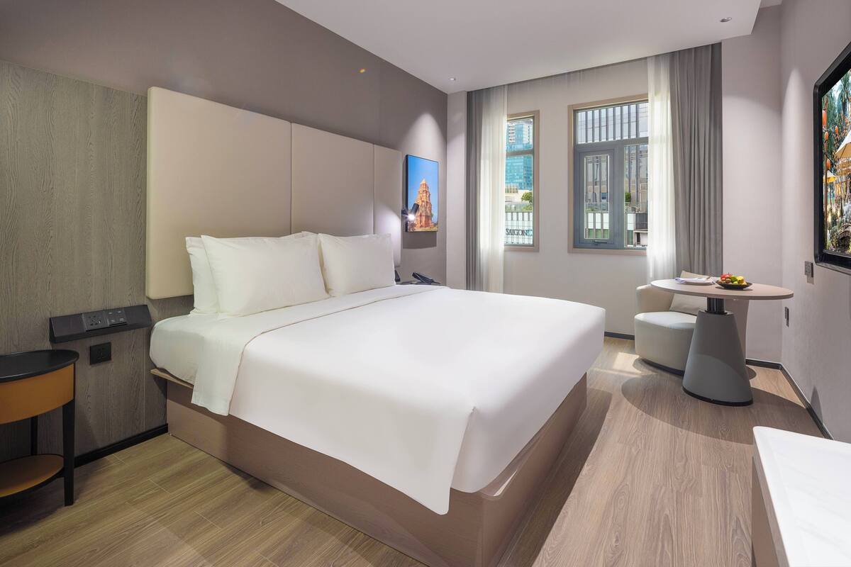 Quarto de hotel com cama grande, mesa de cabeceira, pequena mesa redonda e janela com cortinas.