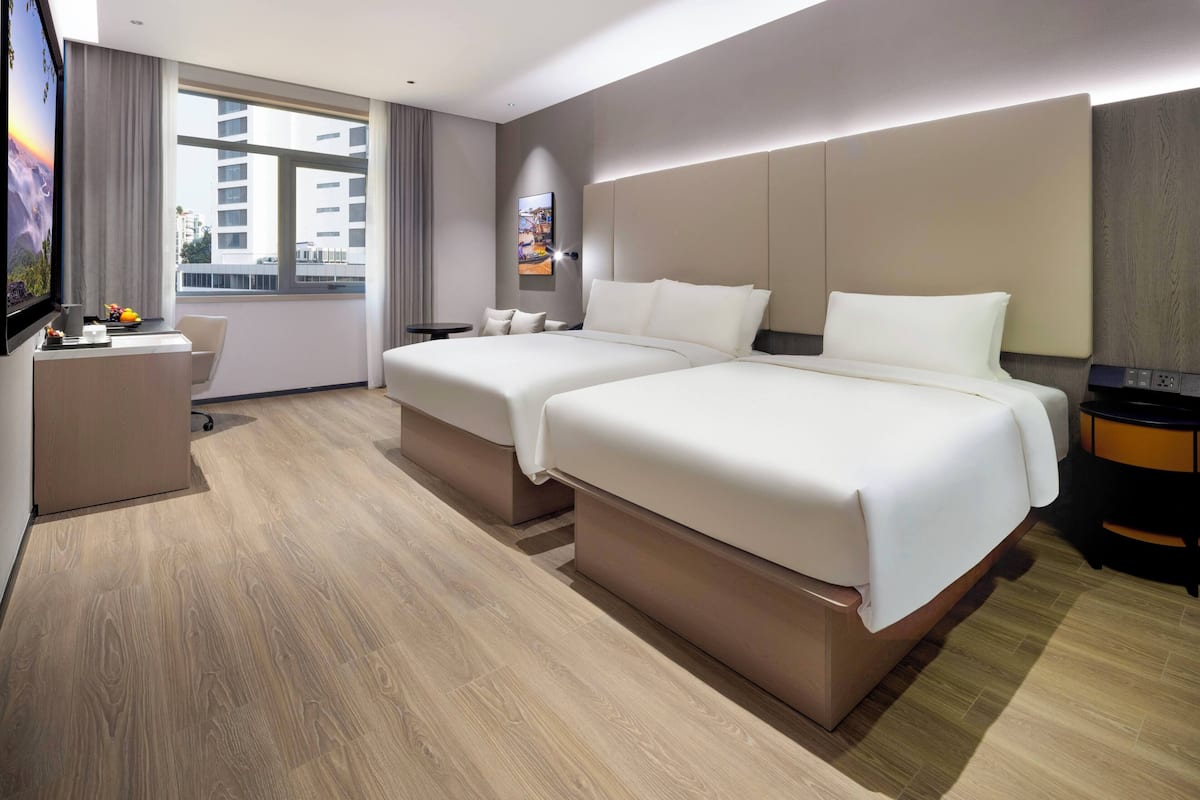 Quarto de hotel com duas camas, uma mesa, uma cadeira, uma televisão e uma janela com cortinas.