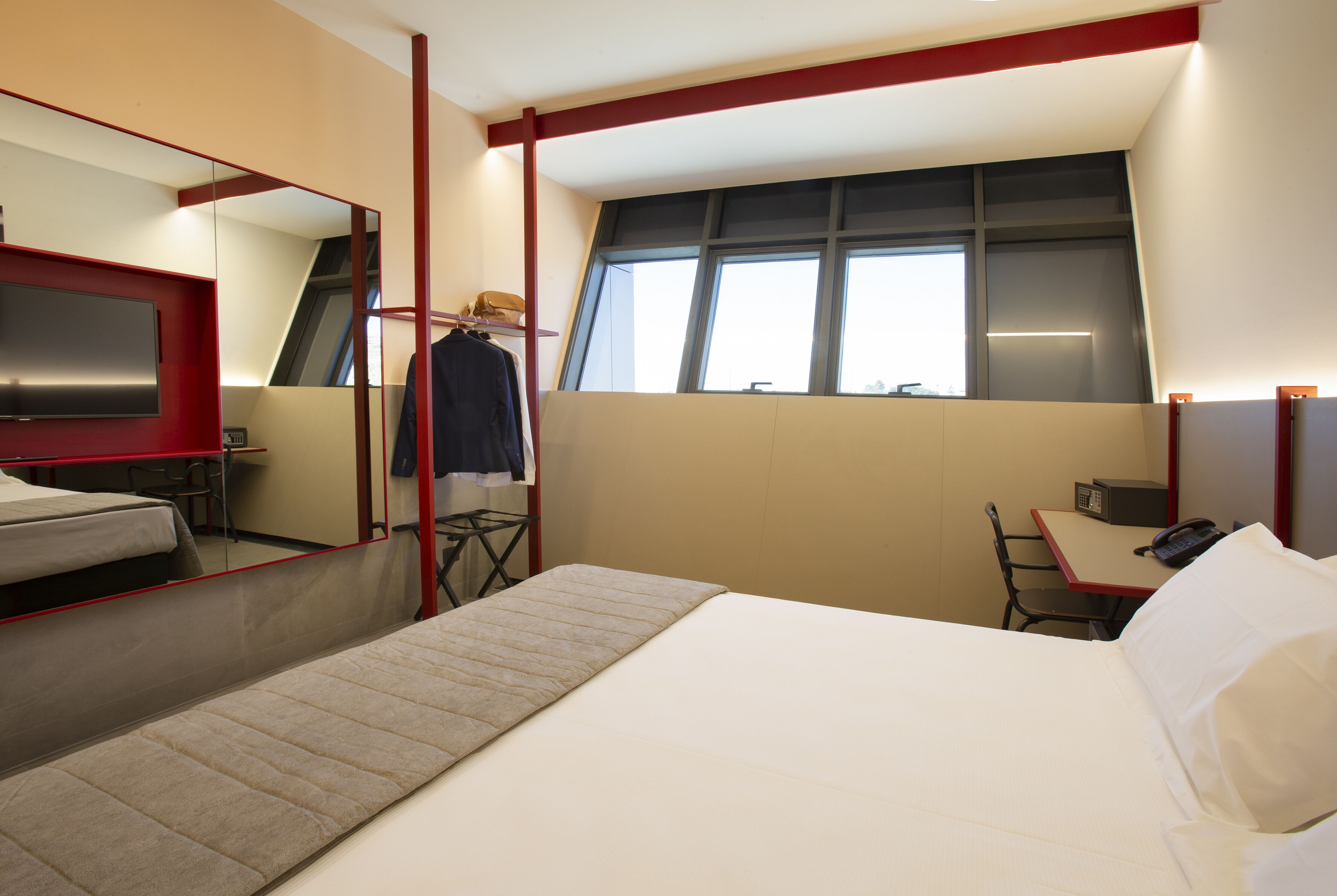 Quarto de hotel com cama, secretária, cadeira, televisão e janela.