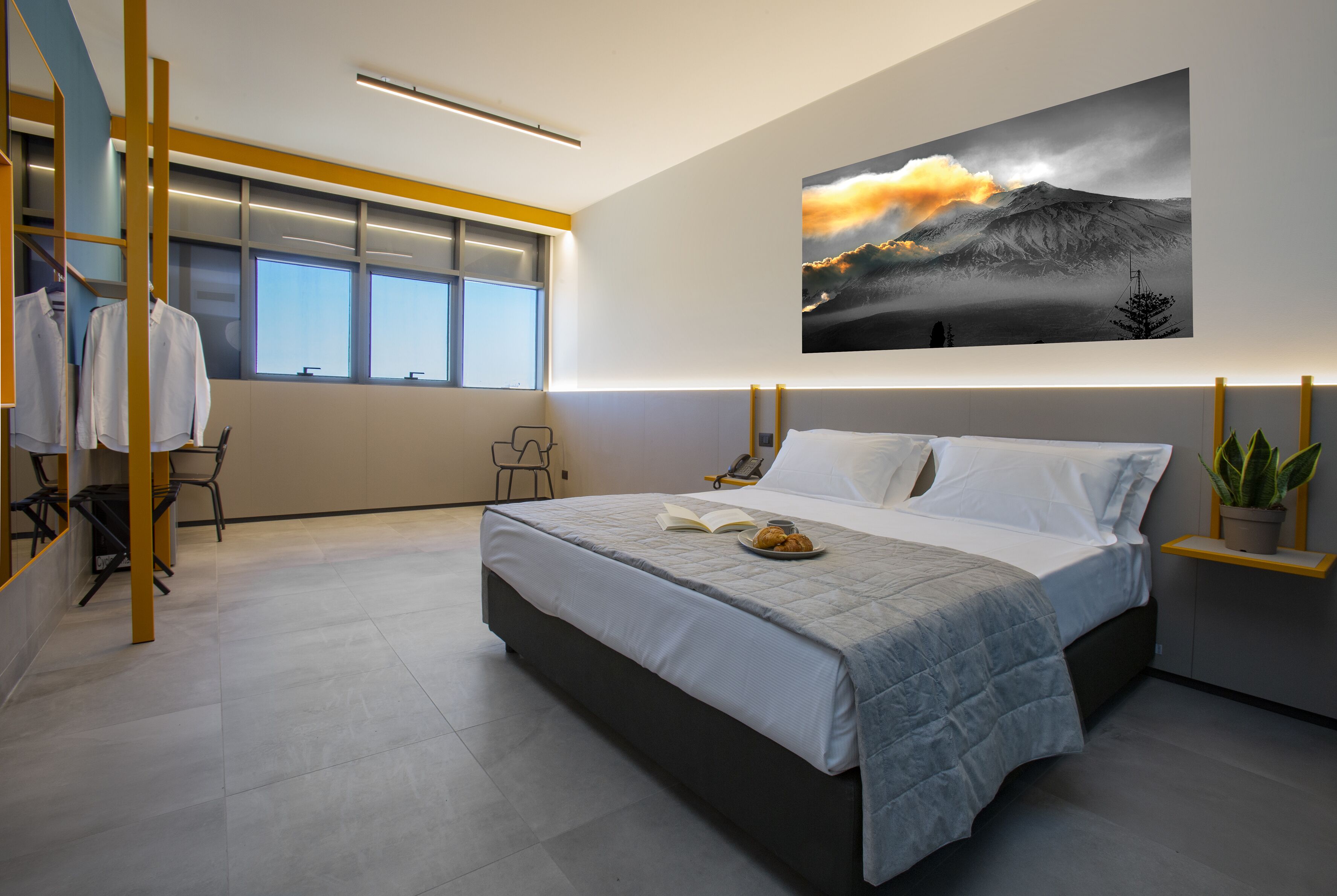 Quarto moderno com uma cama grande, uma janela com vista, um quadro na parede e uma pequena mesa com uma planta.