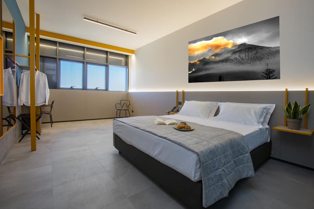 Quarto moderno com uma cama grande, uma janela com vista, um quadro na parede e uma pequena mesa com uma planta.