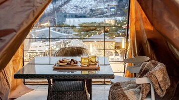 Ambiente acolhedor em interior, com mesa posta para dois, um prato de biscoitos e duas xícaras de chá, uma cadeira de vime e vista pela janela.