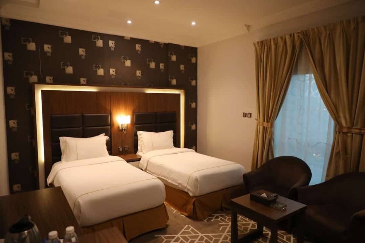 Quarto de hotel com duas camas, uma escrivaninha, uma cadeira e uma janela grande com cortinas.
