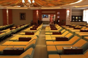 Uma sala de conferências de hotel com fileiras de cadeiras e mesas, um palco e um lustre.