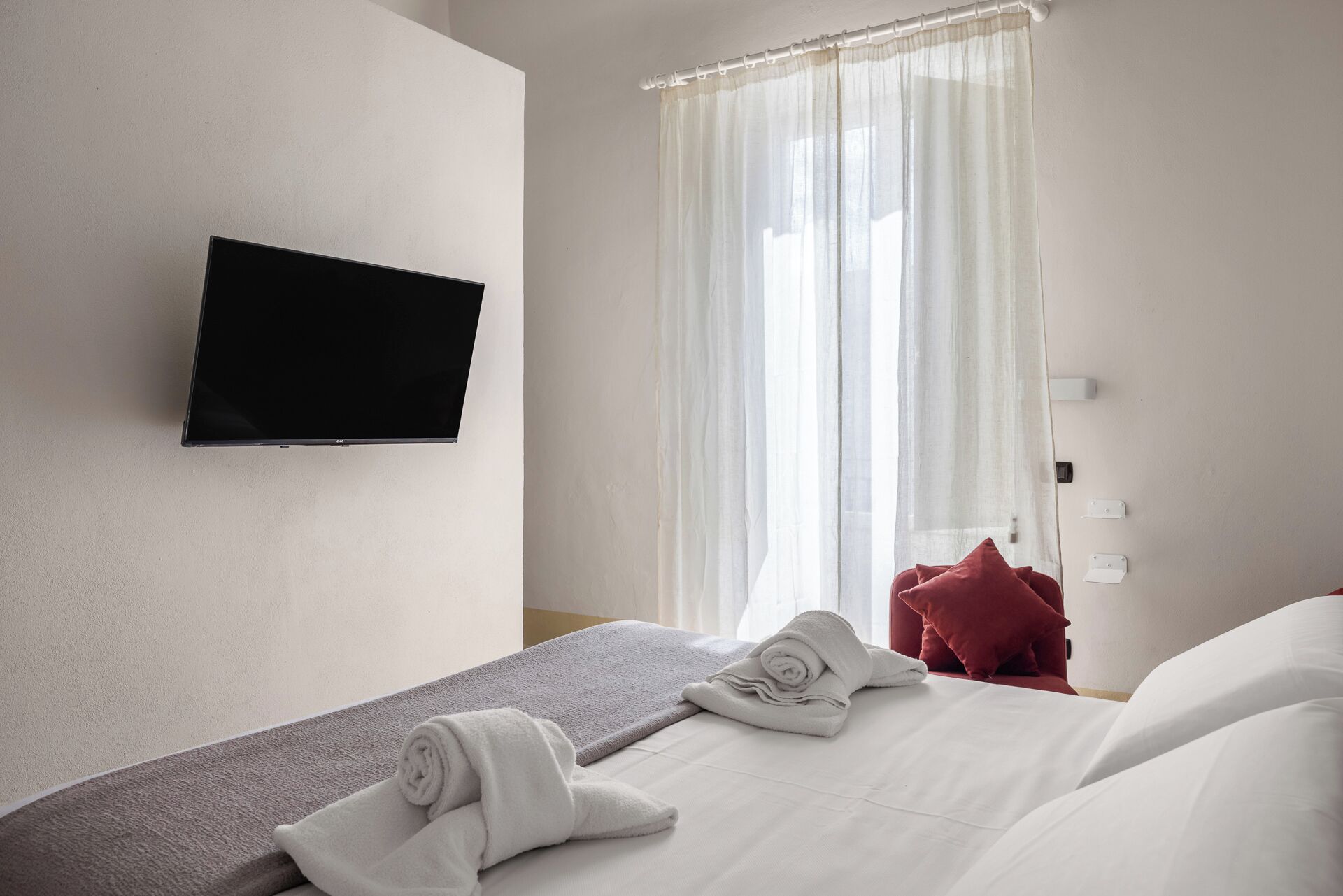 Quarto de hotel com televisão de tela plana na parede, cama com roupa de cama branca, manta cinza e toalhas enroladas.