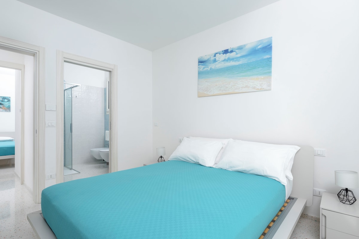 Quarto com cama, banheiro com chuveiro e pintura de uma cena de praia.