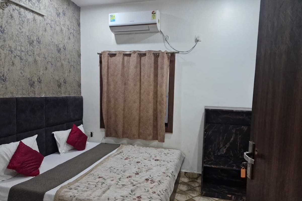 Quarto com cama, cabeceira, janela com cortinas, ar condicionado e porta.